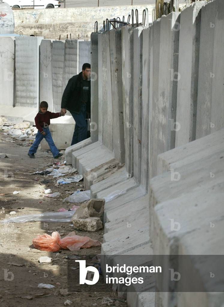 Image of PALESTINIAN WALK ALONG A CONCRETE WALL TO THE KUPSA CHECKPOINT