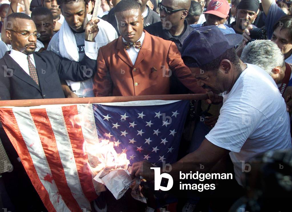 Image of Black Panther Quanell X (C) stands behind a burning U.