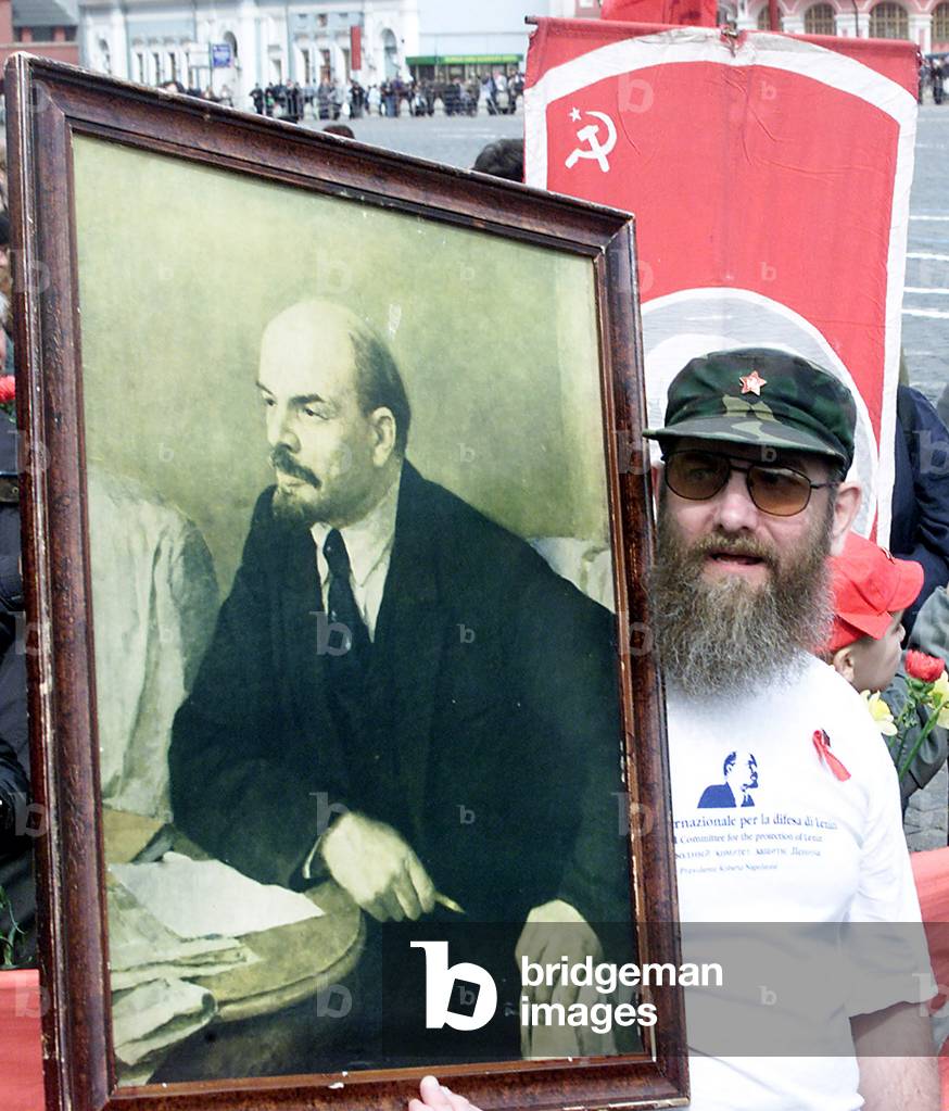 Image of RUSSIAN COMMUNIST STANDS WITH LENIN'S PORTRAIT IN FRONT OF LENIN'S