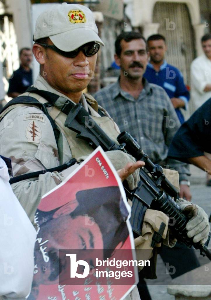 Image of A US SOLDIER STANDS GUARD AS IRAQI POLICEMAN DISTRIBUTES WANTED