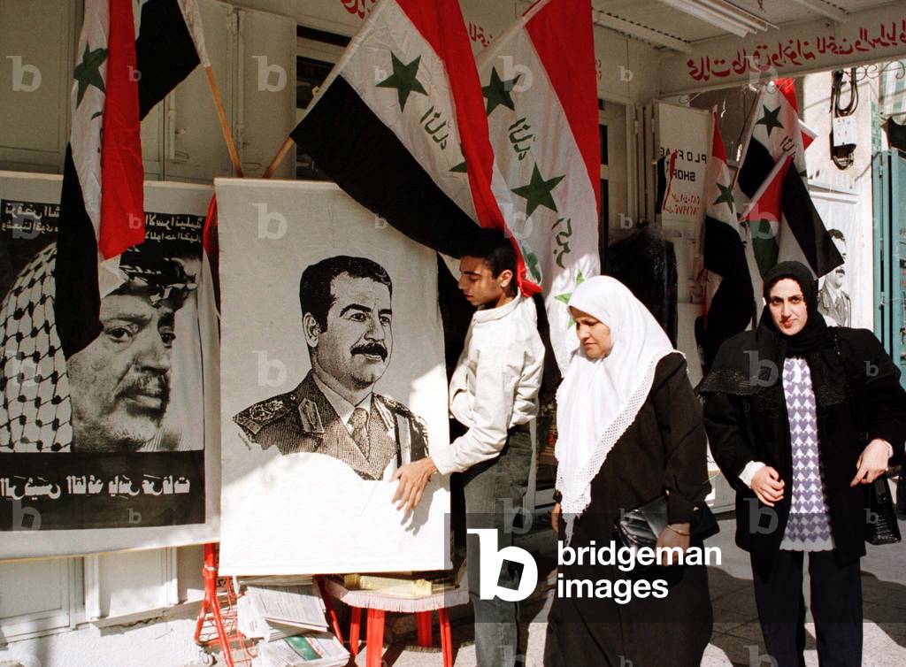 Image of Palestinian women pass a shop selling President Saddam Hussein and