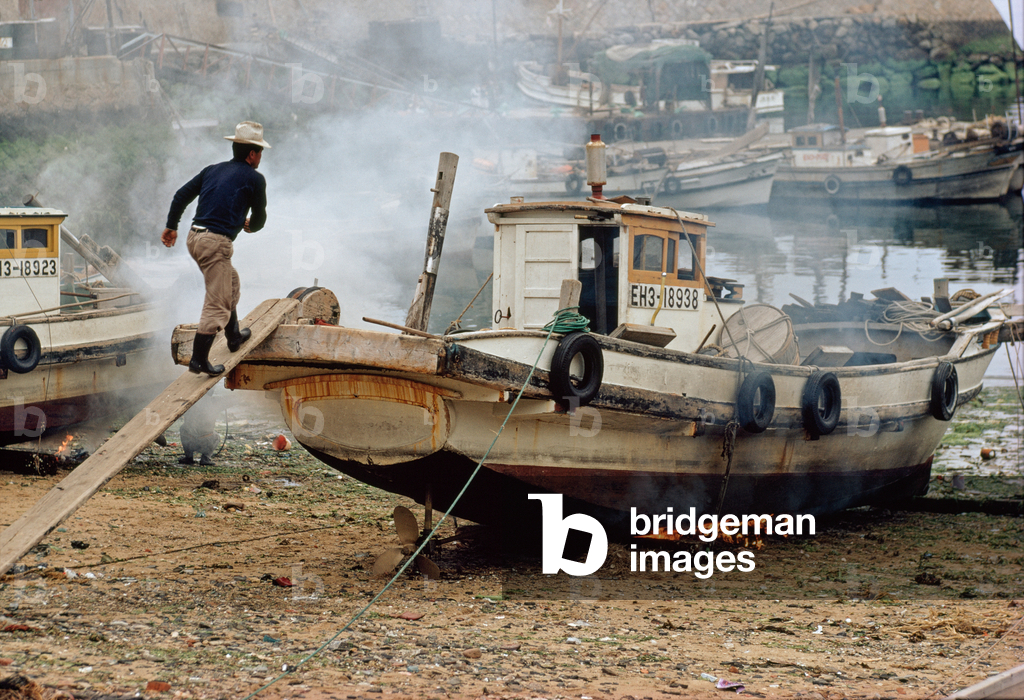 Image of A fisherman scorching his boat hull to kill marine borers. by ...